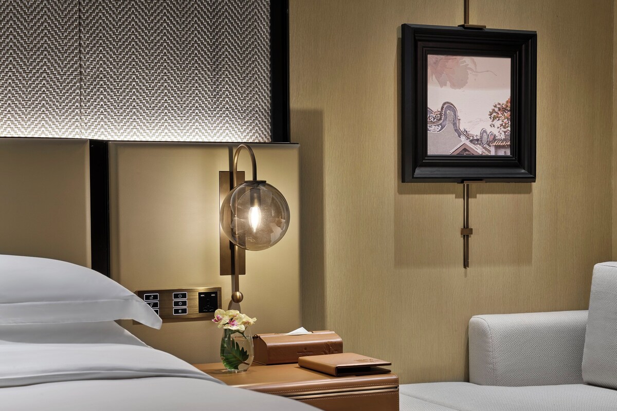 Quarto de hotel com cama, mesa de cabeceira com um vaso de flores, quadro na parede e uma luminária moderna.