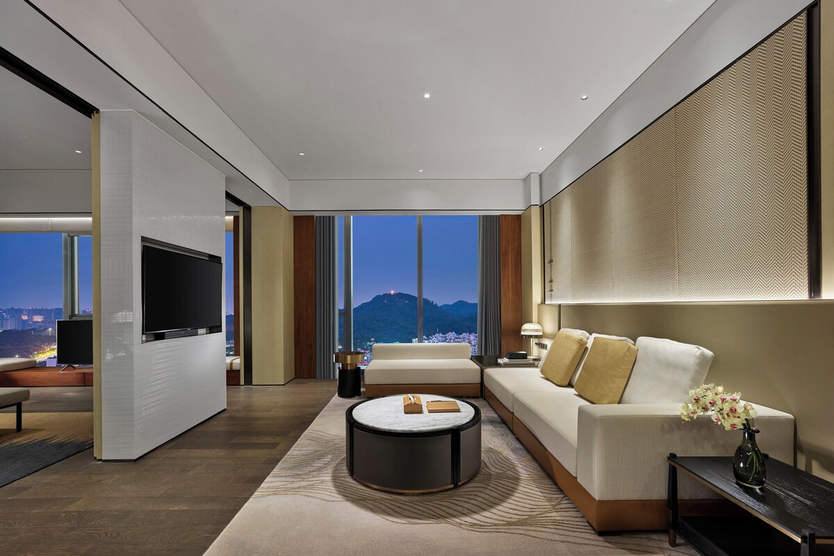Uma sala de estar moderna com uma televisão grande, um sofá, uma mesa de centro e vista para a paisagem urbana ao entardecer.