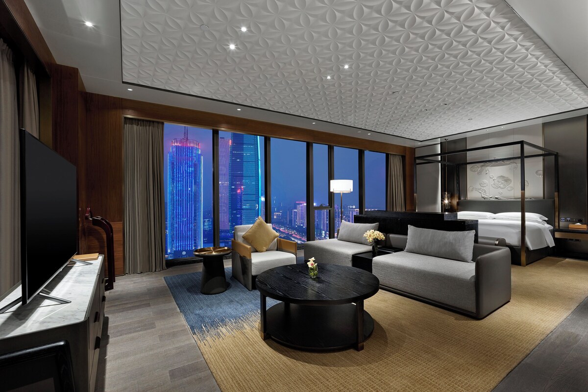 Quarto de hotel moderno com vista para a cidade, uma cama grande, um sofá, uma mesa de centro e uma televisão.