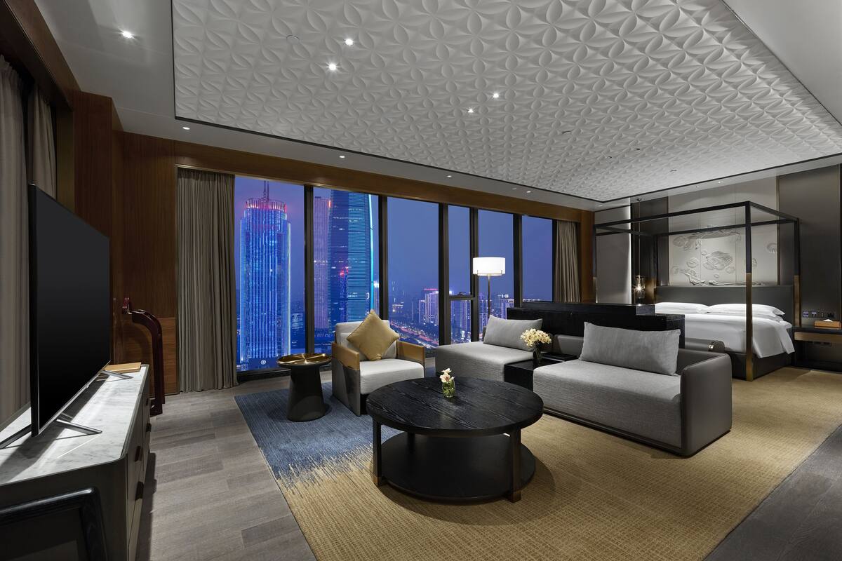 Quarto de hotel moderno com vista para a cidade, uma cama grande, um sofá, uma mesa de centro e uma televisão.