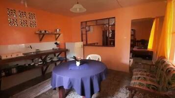 Um espaço compacto com área de refeições, incluindo uma mesa redonda e cadeiras, cozinha compacta com fogão e pia, e sofá com almofadas estampadas.