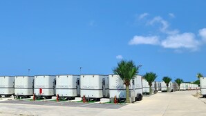 Uma fileira de trailers brancos com a marcação 'TRA+CAN', estacionados sobre uma superfície de concreto, com palmeiras e um céu azul claro ao fundo.