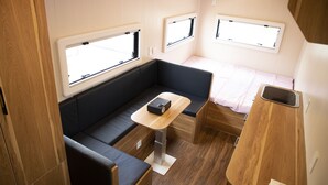 Interior compacto de um veículo recreativo, com cozinha compacta, área de estar e duas janelas.