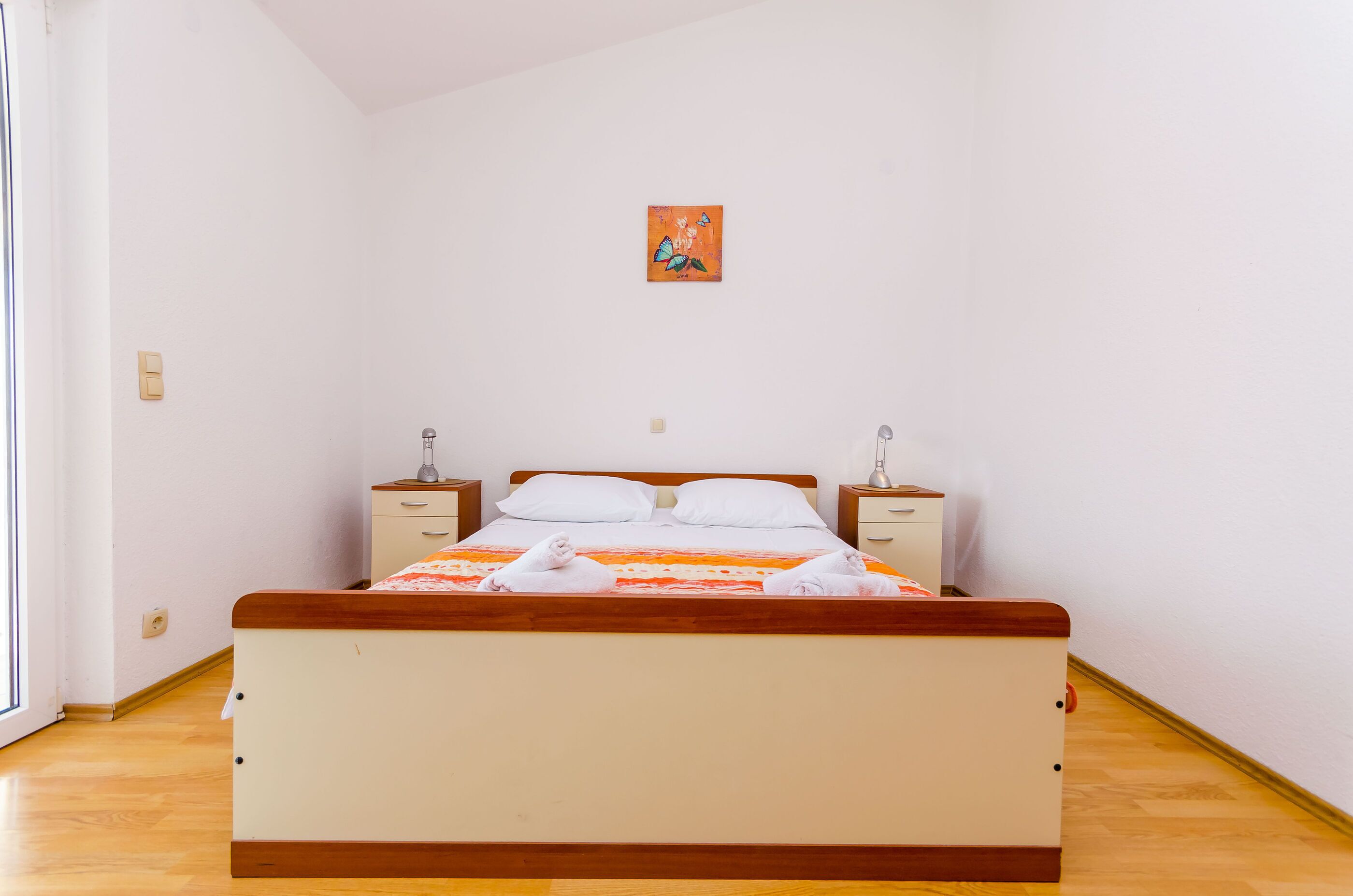 Quarto com cama de madeira, almofadas brancas, um pequeno quadro na parede e uma janela à esquerda.