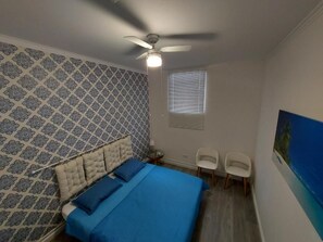 Quarto com cama, ventilador de teto, janela com persianas e um quadro na parede.