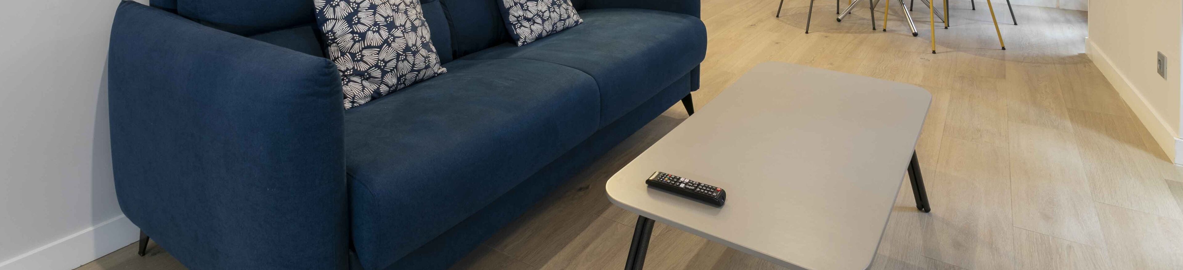 Uma sala de estar com um sofá azul, almofadas estampadas e um controle remoto sobre uma mesa de centro branca.