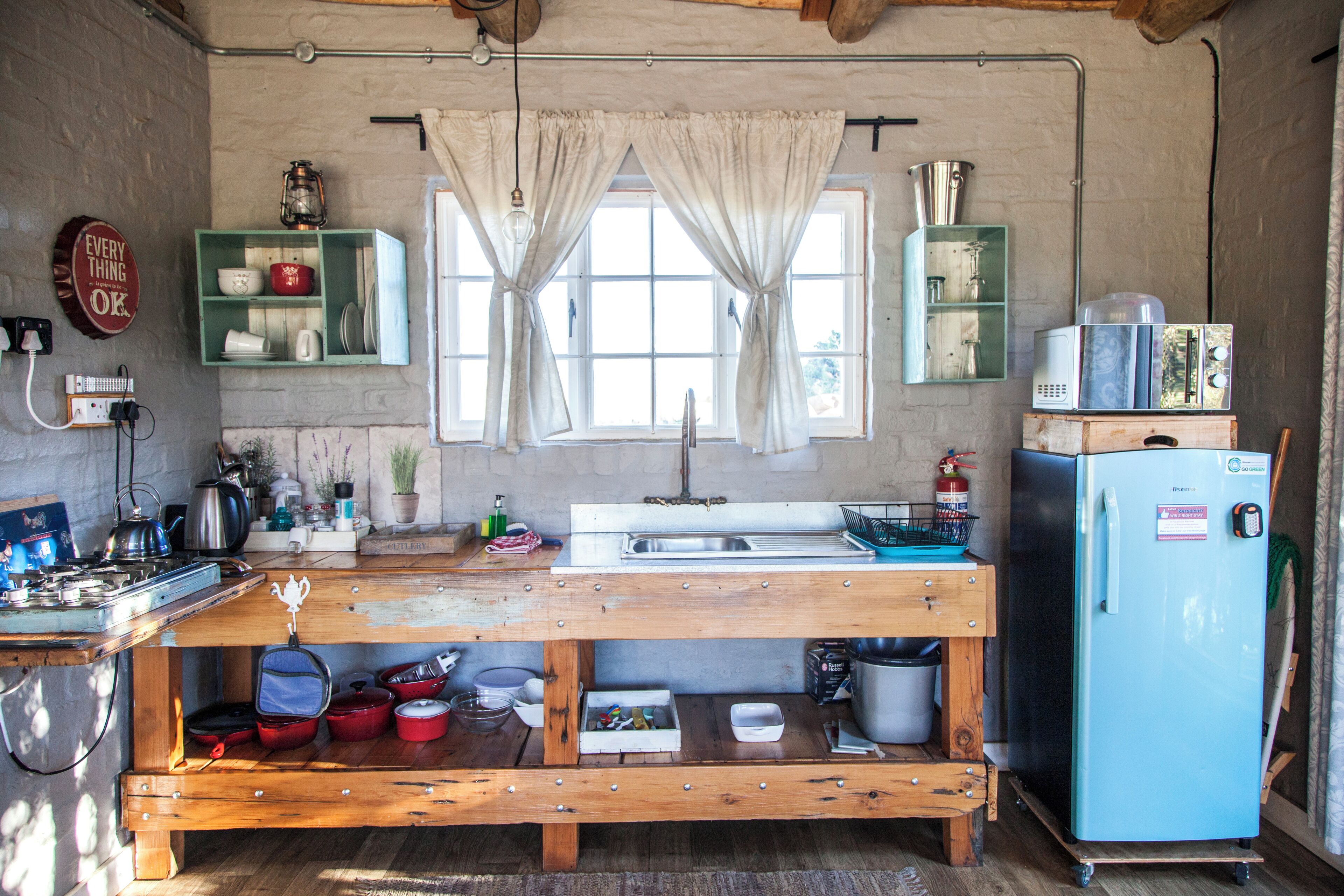 Uma cozinha rústica com bancada de madeira, pia, janela com cortinas e uma geladeira azul.