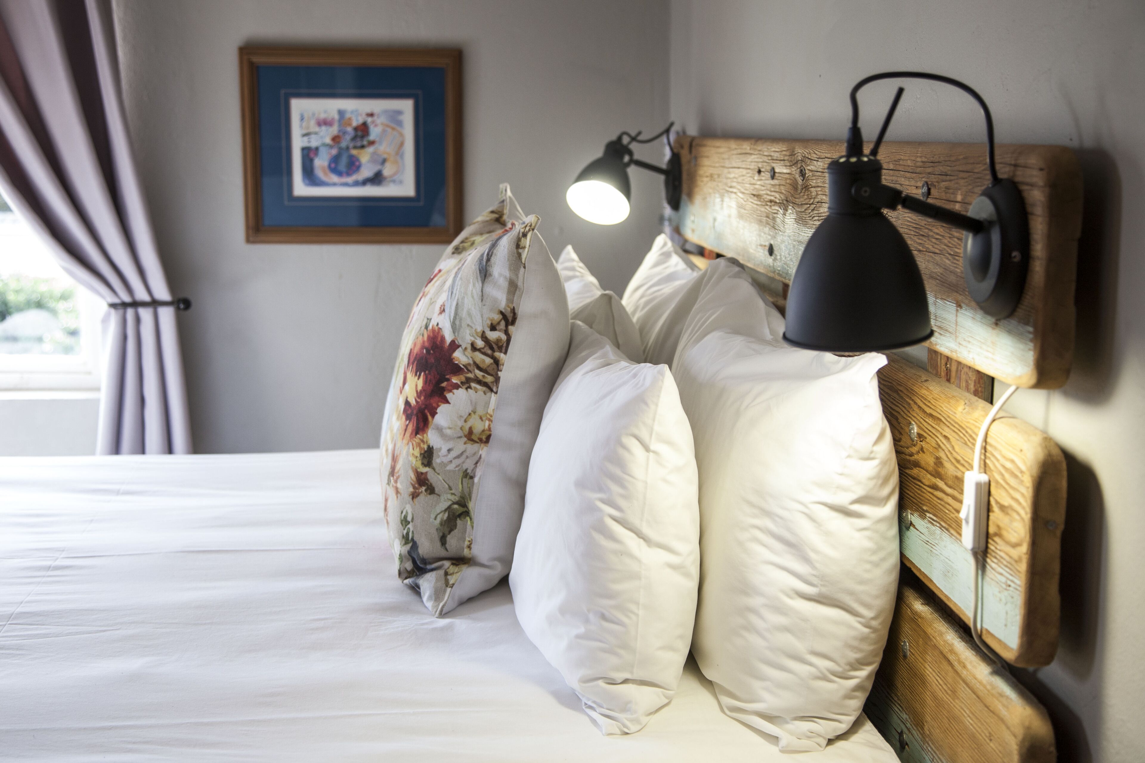 Cama com almofadas brancas e florais, cabeceira de madeira e luminária fixada na parede.
