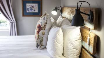 Cama com almofadas brancas e florais, cabeceira de madeira e luminária fixada na parede.