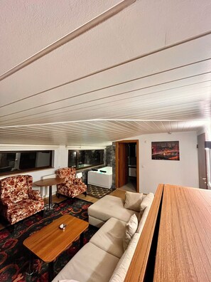 Uma sala de estar moderna com sofá, poltronas, mesa de centro e mesa de madeira. Há uma janela com vista para a cidade à noite.