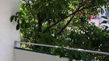 Varanda com guarda-corrimão de metal, chão de azulejos e vista para vegetação verde e um edifício ao fundo.