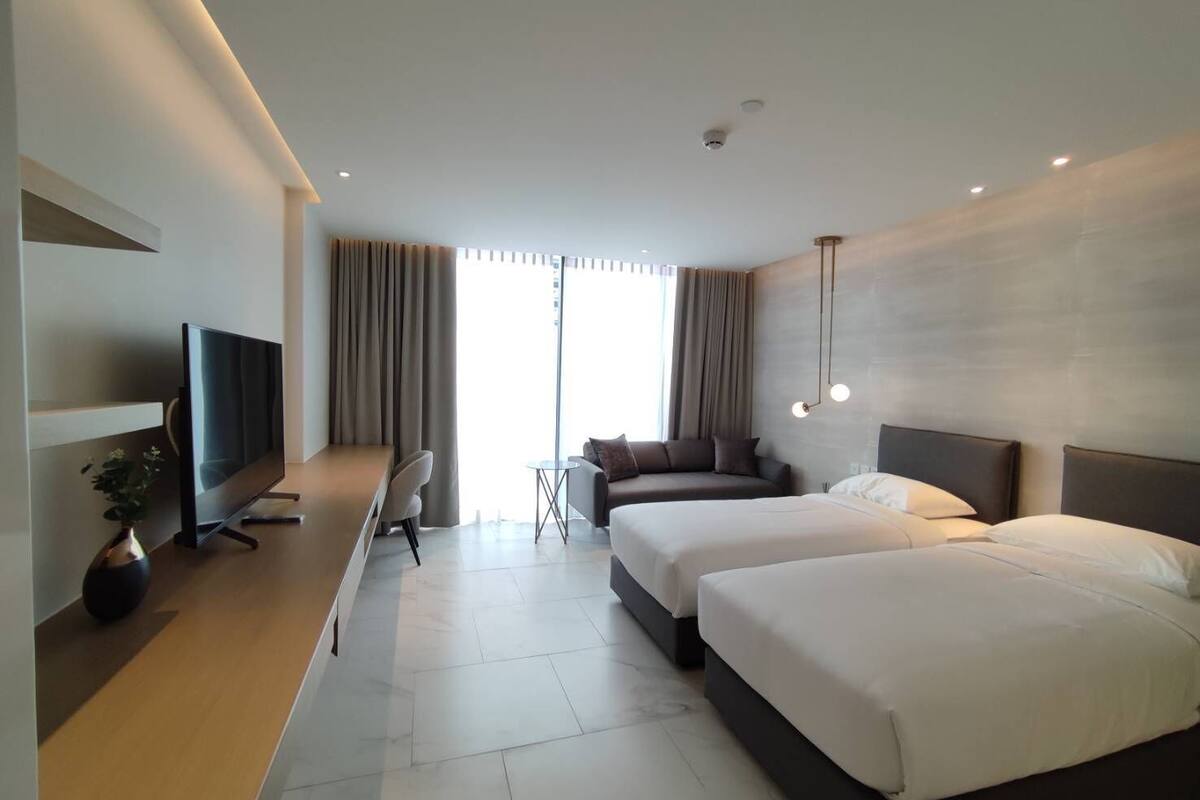 Quarto de hotel moderno com duas camas, televisão de tela plana, área de estar e janelas amplas.