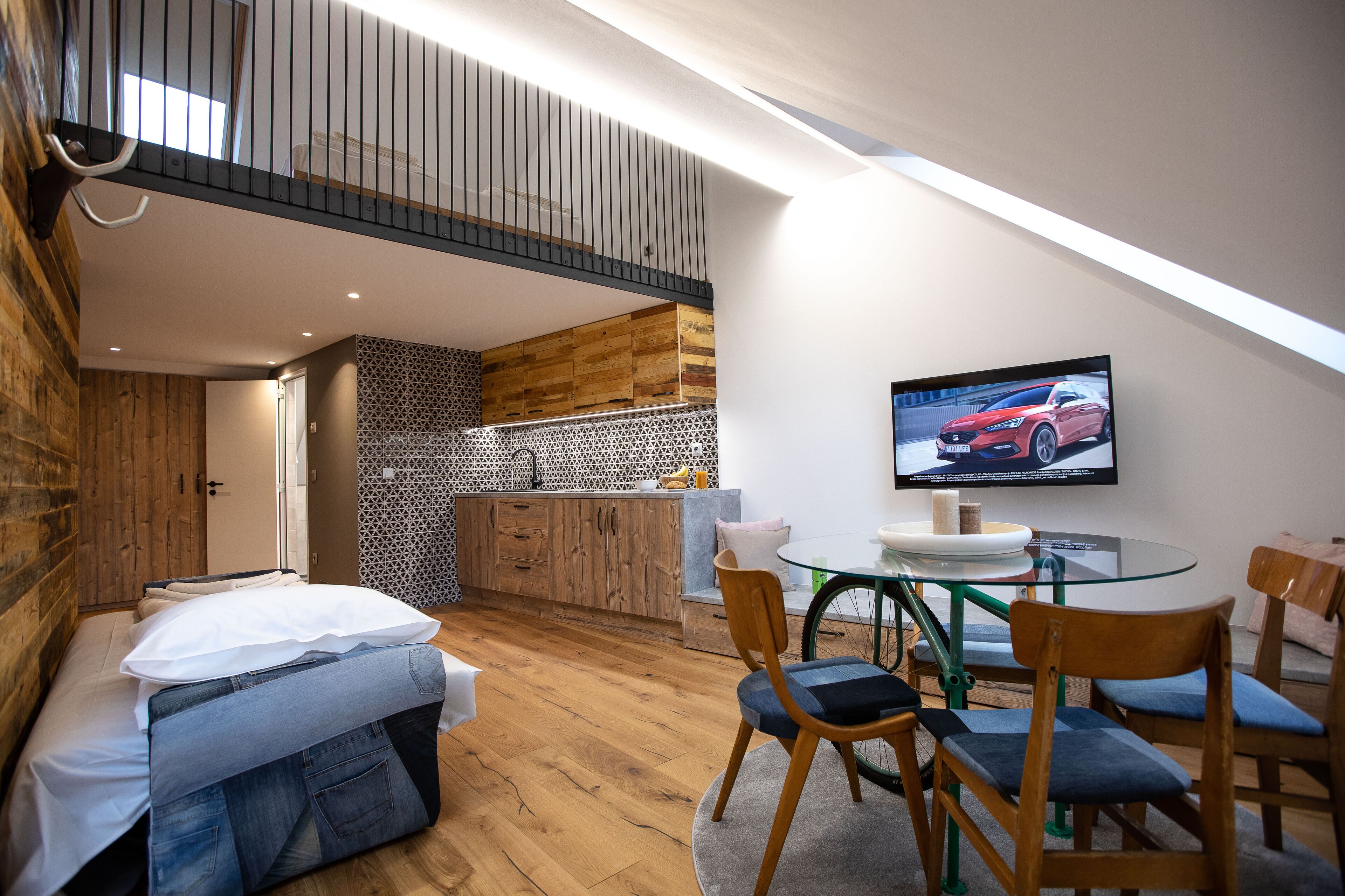 Quarto estilo loft moderno com cama mezanina, área de refeições com mesa de vidro e cozinha com armários de madeira.