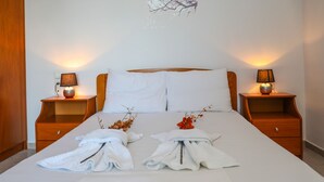 Cama bem arrumada com lençóis e toalhas brancas, duas mesas de cabeceira com abajures, e uma decoração na parede com uma árvore e pássaros.