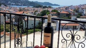 Uma mesa com uma garrafa de vinho e uma tigela de frutas numa varanda com vista para uma cidade costeira.