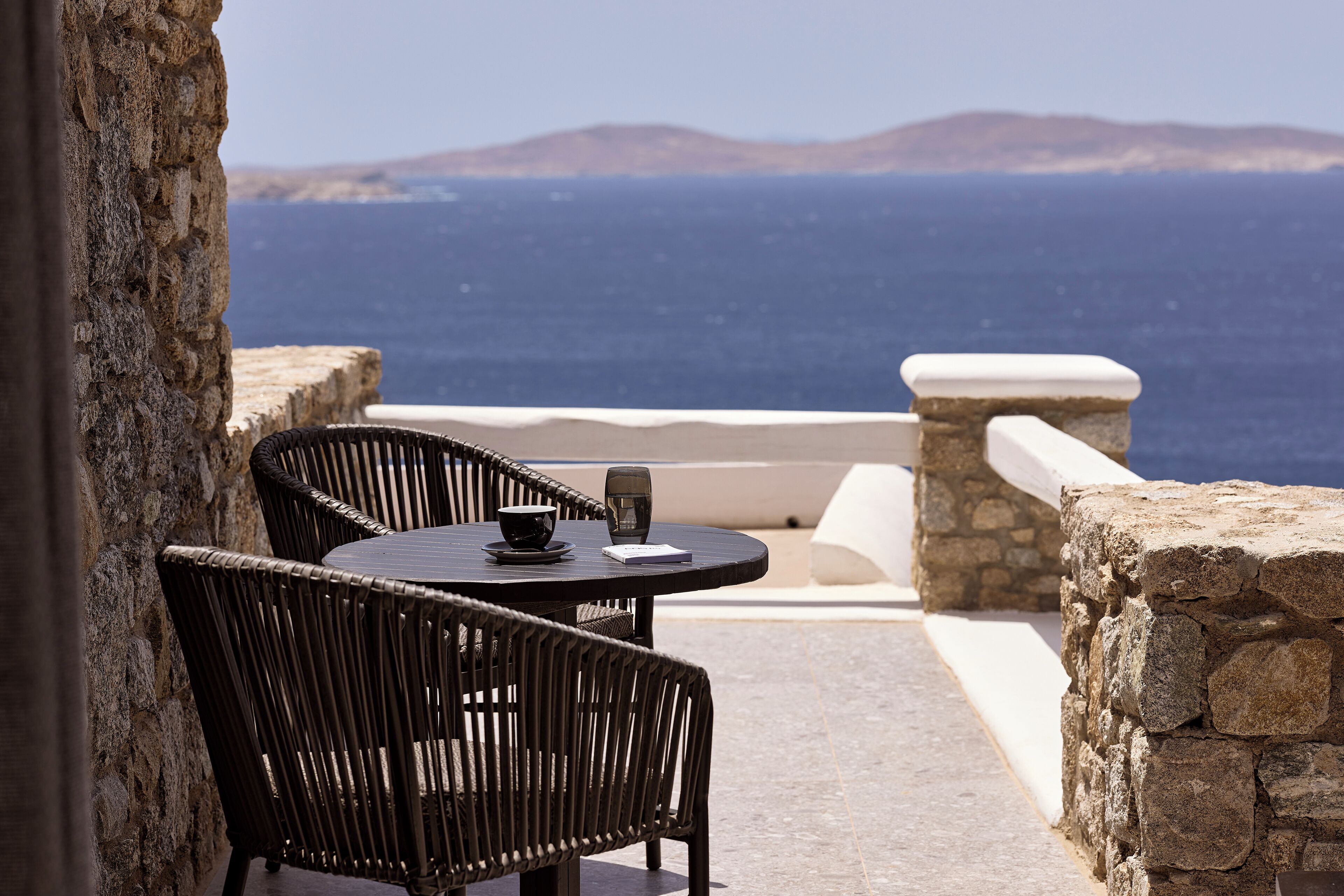 Terraço com cadeiras de vime e uma mesa, com vista para o mar e montanhas ao longe.