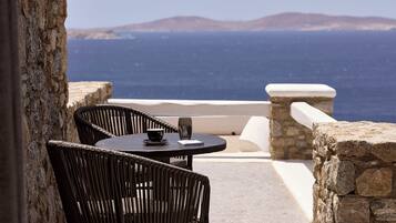 Terraço com cadeiras de vime e uma mesa, com vista para o mar e montanhas ao longe.