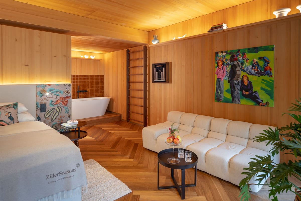 Quarto de hotel com decoração em madeira, um sofá, uma mesinha com um vaso e frutas, uma banheira e um quadro na parede.