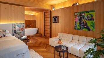 Quarto de hotel com decoração em madeira, um sofá, uma mesinha com um vaso e frutas, uma banheira e um quadro na parede.