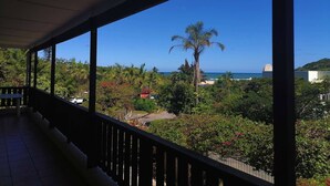 Varanda coberta com vista para uma paisagem tropical, incluindo palmeiras e uma praia.