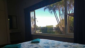 Quarto de hotel com uma janela grande, cama com roupa de cama floral e vista para palmeiras e uma cerca lá fora.