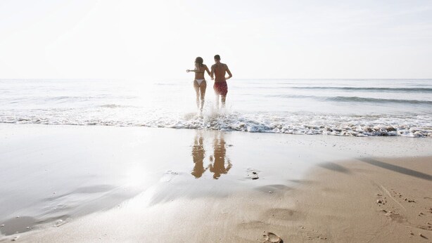 Duas pessoas caminham numa praia de areia, com ondas ao fundo.