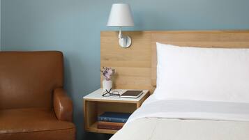 Quarto de hotel com poltrona de couro marrom, mesa de cabeceira de madeira com luminária branca, um vaso com flores e cama com roupa de cama branca.