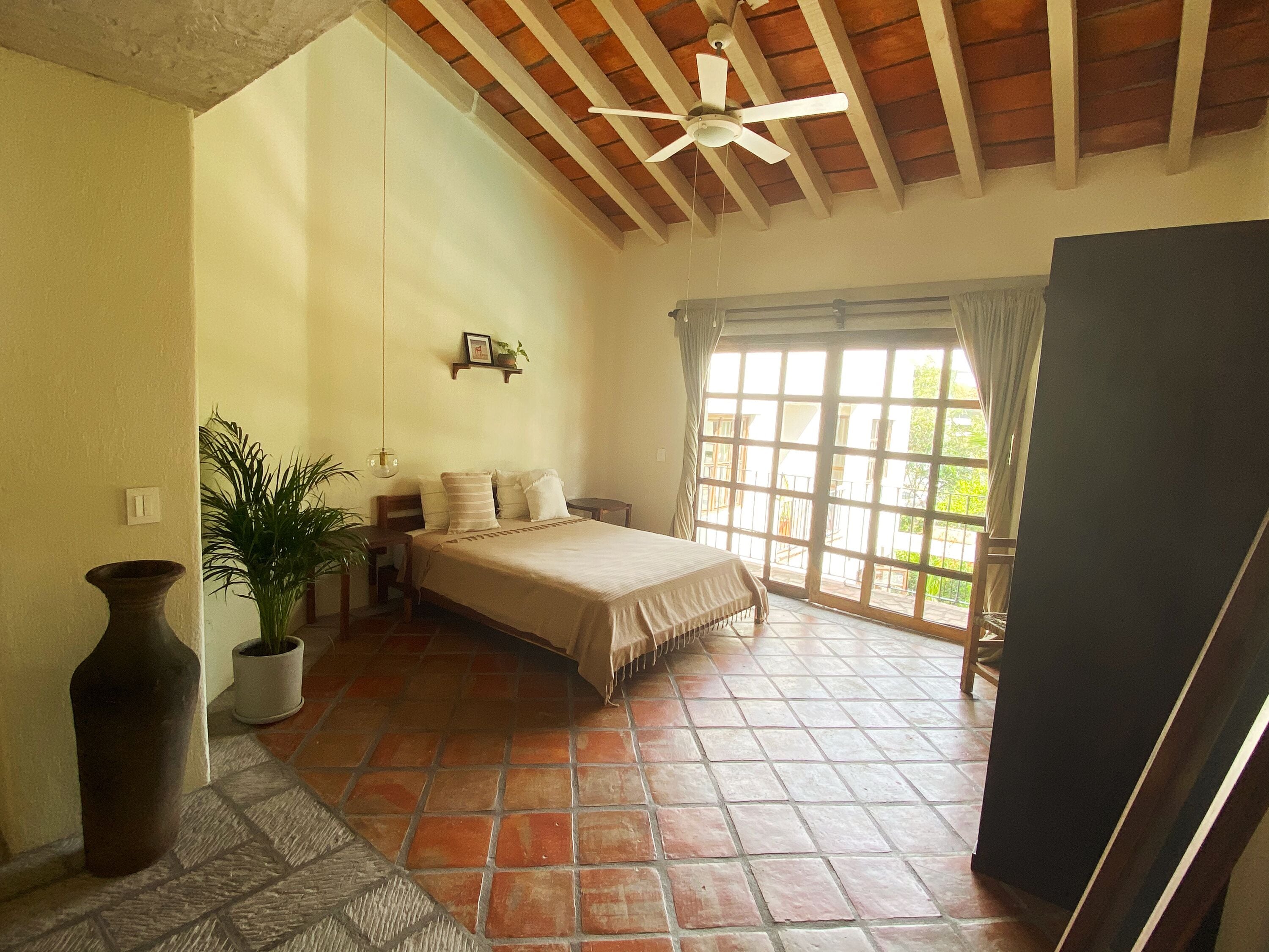 Quarto com cama, planta em vaso, ventoinha no teto e janela com cortinas.