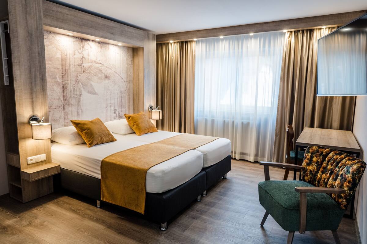 Quarto de hotel moderno com uma cama grande, uma mesa de cabeceira com abajur, uma cadeira e uma janela ampla com cortinas.