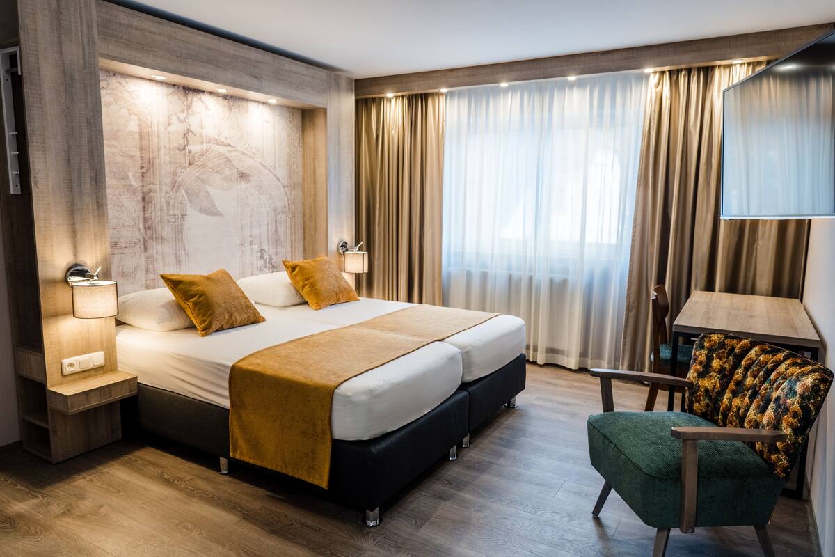 Quarto de hotel moderno com uma cama grande, uma mesa de cabeceira com abajur, uma cadeira e uma janela ampla com cortinas.
