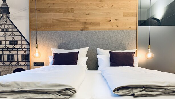 Uma cama de casal com lençóis brancos, duas almofadas escuras e cabeceira de madeira.