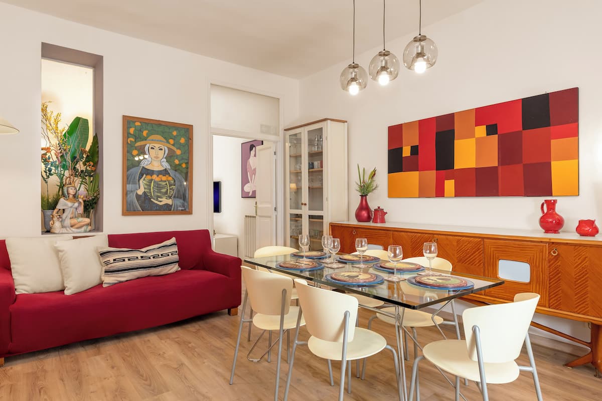 Uma sala de estar moderna com um sofá vermelho, mesa de jantar de vidro e cadeiras brancas. Há objetos decorativos nas paredes e um grande quadro abstrato acima da mesa.