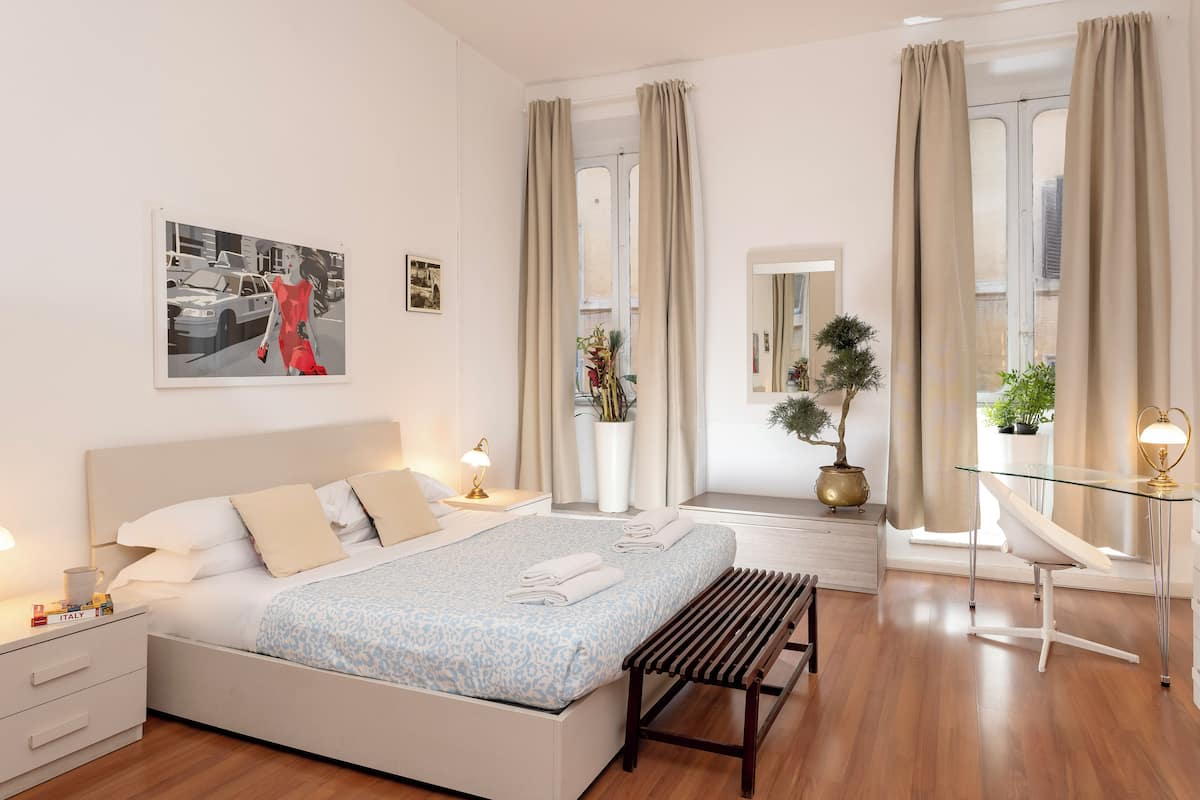 Quarto de hotel moderno com cama, mesas de cabeceira, escrivaninha e uma janela grande.