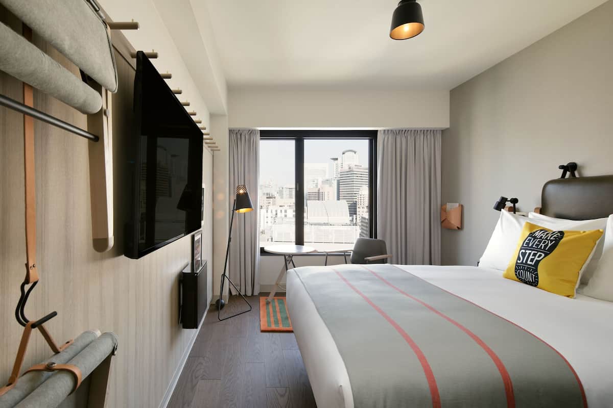 Quarto de hotel com cama, escrivaninha, cadeira, televisão e janela com vista para a cidade.