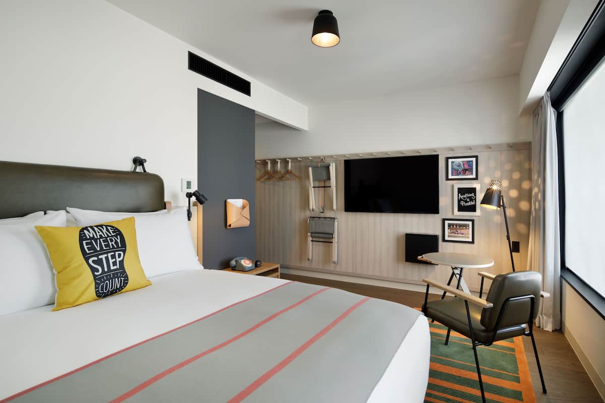 Quarto de hotel com cama, secretária, cadeira, televisão e janela.