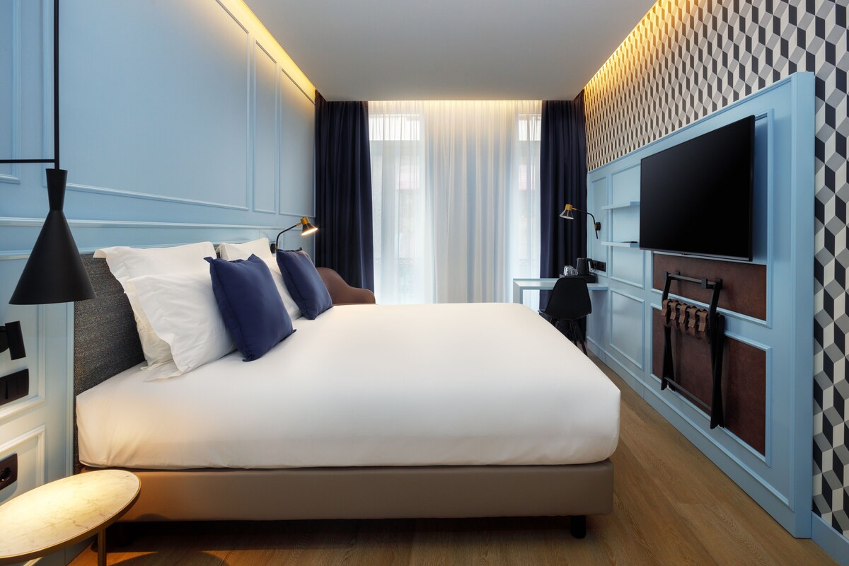 Quarto de hotel moderno com uma cama grande, televisão de tela plana na parede, escrivaninha com cadeira e uma mesinha.