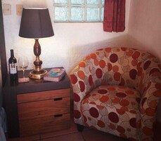 Quarto de canto com cômoda de madeira, luminária, garrafa de vinho e poltrona estampada.