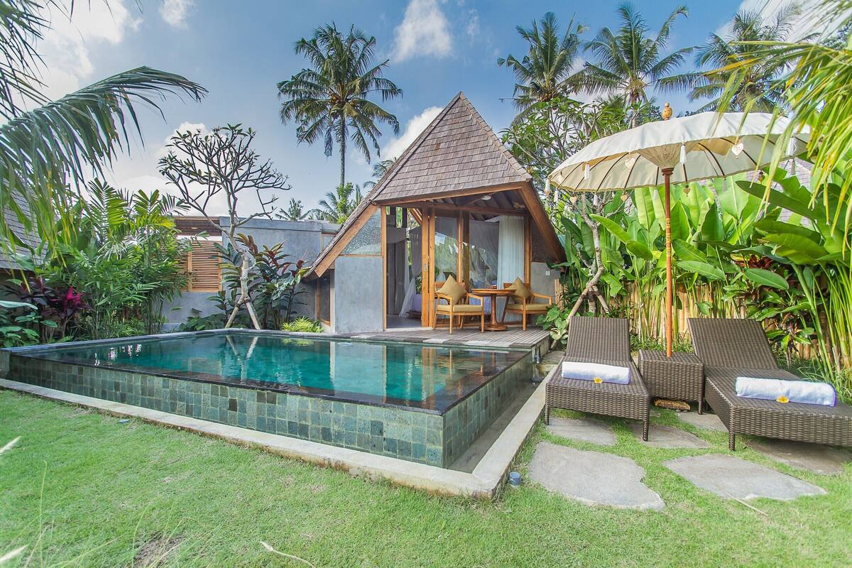 Uma piscina infinita de vidro com um deck de madeira, espreguiçadeiras e um pequeno pavilhão com mesa e cadeiras.