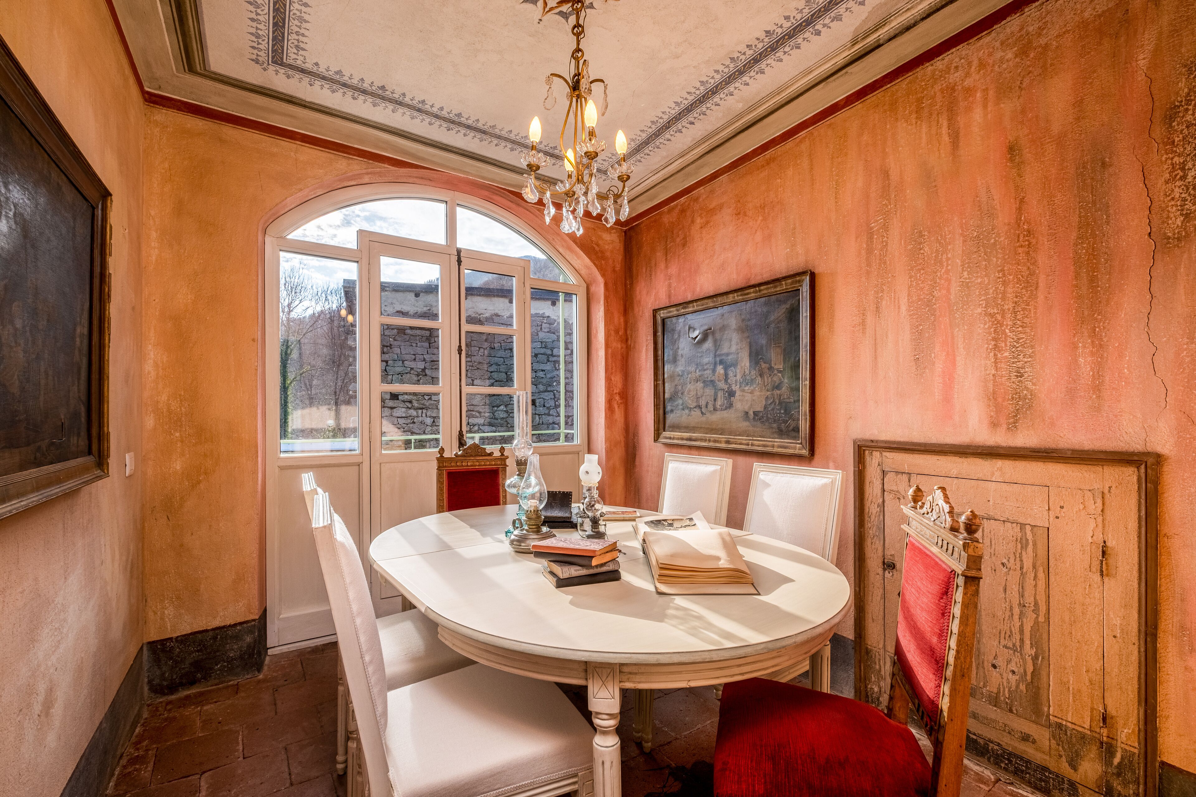 Uma sala de jantar com uma mesa branca, cadeiras e um candelabro. Há uma janela com vista para um edifício de pedra.