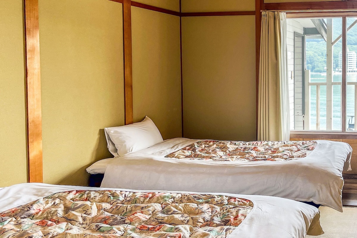 Quarto com duas camas, piso de madeira, janela com vista para um corpo d'água e colinas, e uma porta.