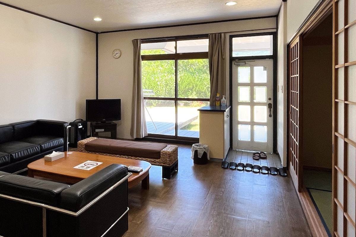 Sala de estar com sofá de couro preto, mesa de centro de madeira, televisão e porta de vidro deslizante para o exterior.