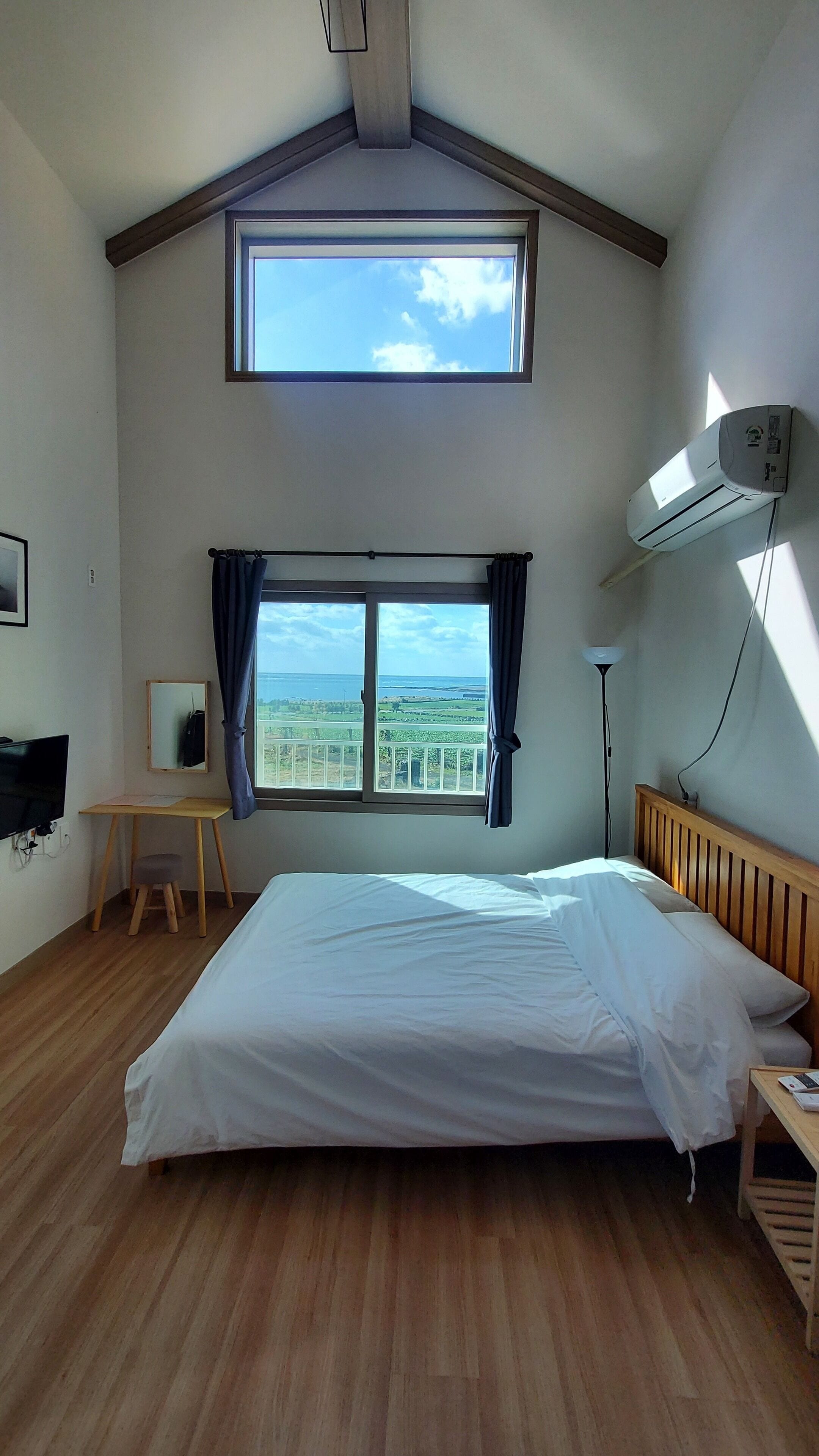 Quarto com uma cama grande, escrivaninha, cadeira, televisão, janela com cortinas e ar condicionado.