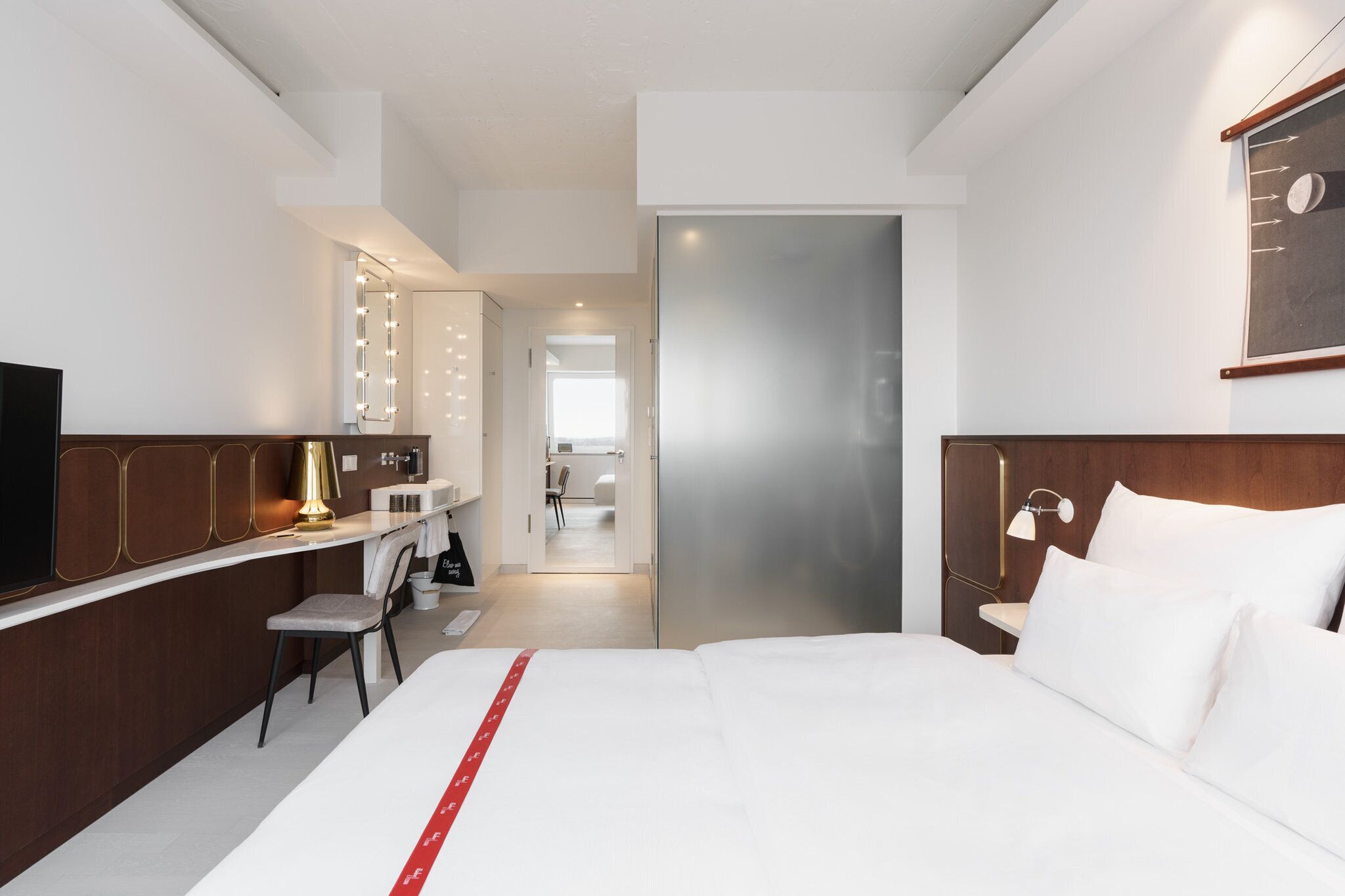 Quarto de hotel com uma cama, uma escrivaninha com cadeira, uma penteadeira iluminada, um espelho e uma porta que dá para outro cômodo.