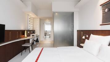 Quarto de hotel com uma cama, uma escrivaninha com cadeira, uma penteadeira iluminada, um espelho e uma porta que dá para outro cômodo.