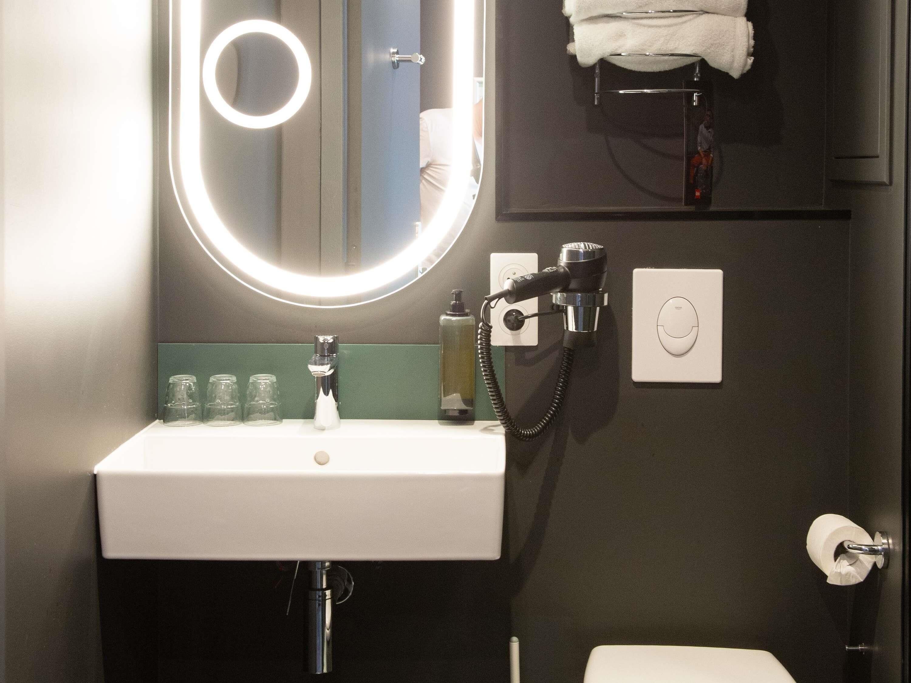 Casa de banho com lavatório branco, espelho com luz, secador de cabelo e um porta-toalhas.