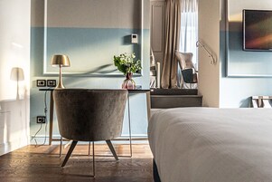 Quarto de hotel com uma cama, uma escrivaninha com abajur, uma cadeira, um vaso com flores e uma janela grande com cortinas.