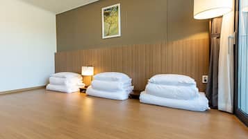 Quarto de hotel com piso de madeira, cama com roupa de cama branca, mesa de cabeceira com luminária e janela com cortinas.