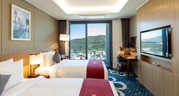 Quarto de hotel com duas camas, uma escrivaninha, televisão e uma janela grande com vista para a cidade.