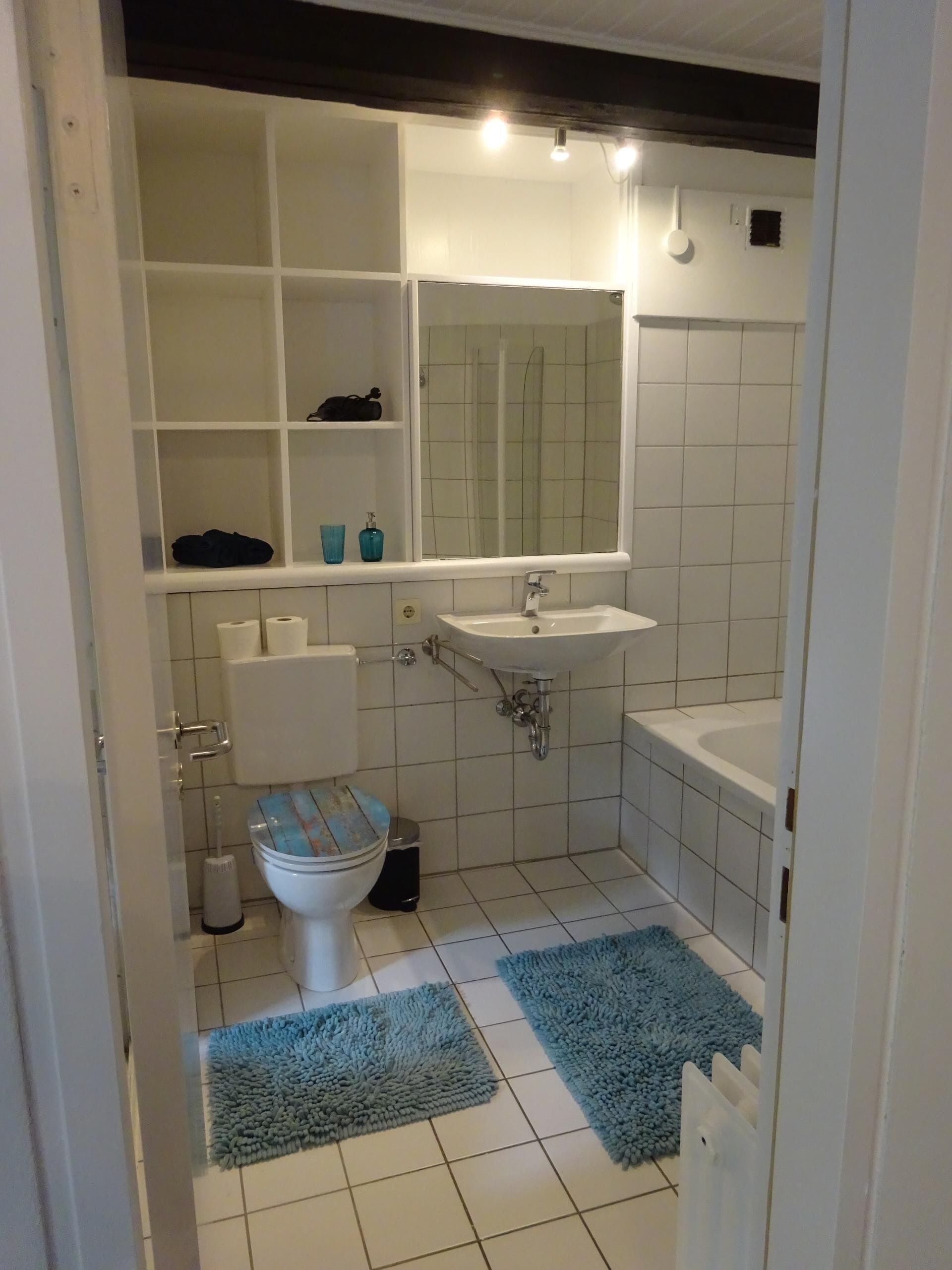 Casa de banho com sanita, lavatório e banheira. Existem tapetes azuis no chão e uma prateleira com artigos de higiene pessoal.