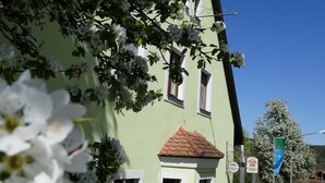 Um edifício de dois andares com fachada verde e telhado de telhas vermelhas. Há uma placa com um logotipo e texto no lado direito do edifício.
